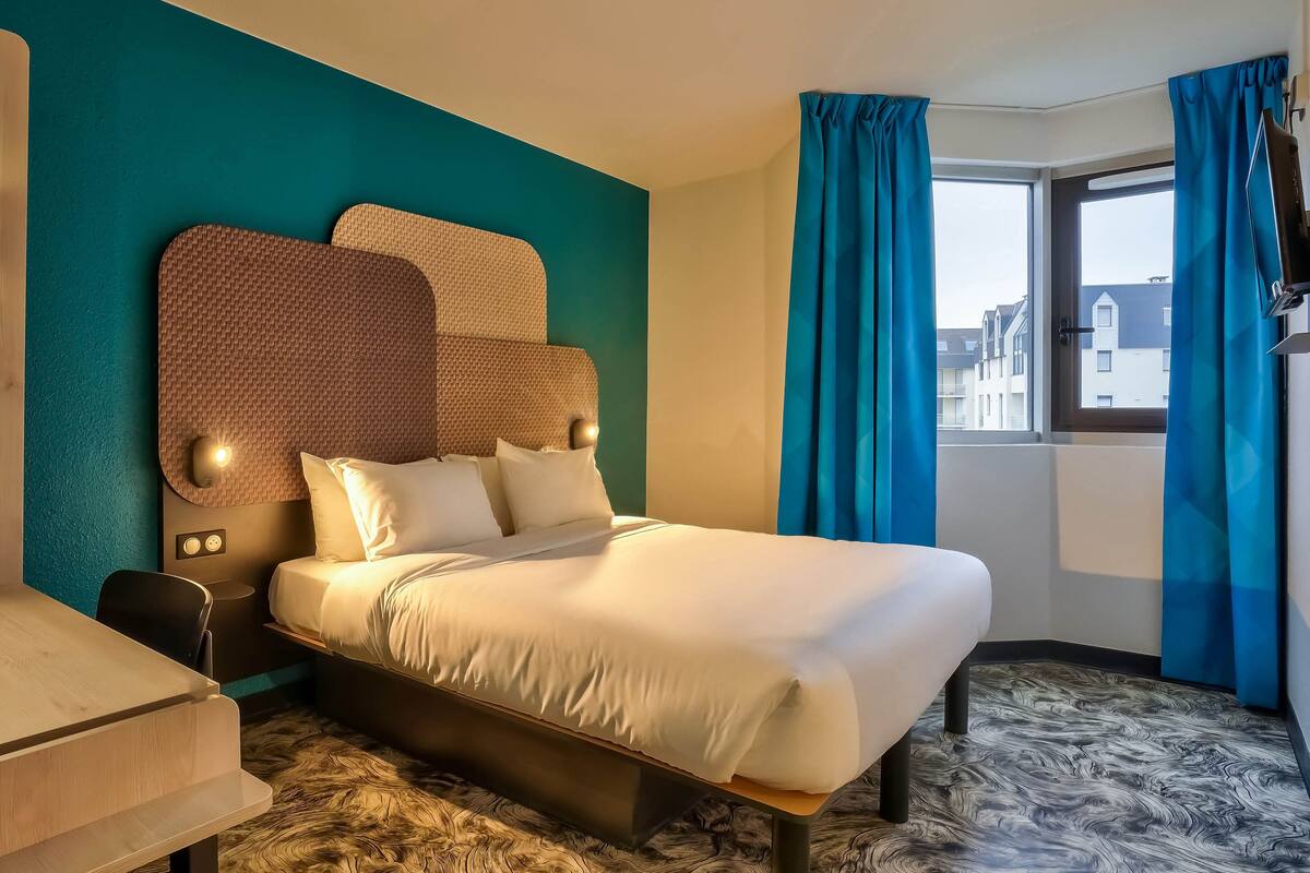 Quarto de hotel com cama, escrivaninha e vista para prédios pela janela.