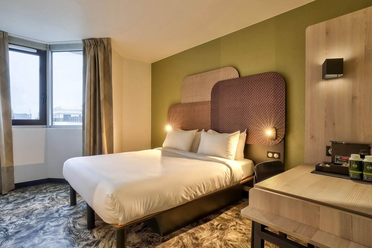 Quarto de hotel com uma cama grande, uma escrivaninha com cafeteira e vista para a cidade pela janela.