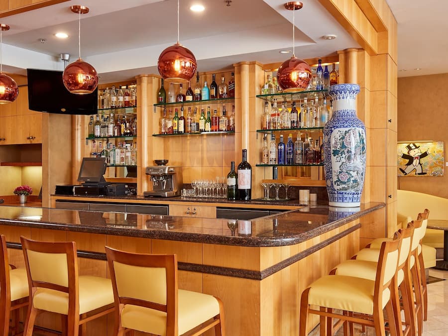 Uma área de bar com balcão de madeira, diversas opções de assento e variedade de garrafas e copos nas prateleiras.