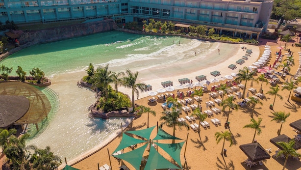 Vista aérea de um resort com uma piscina grande, área de praia com espreguiçadeiras e um prédio de hotel ao fundo.