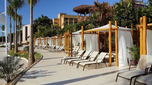 Uma área de piscina em resort, com espreguiçadeiras, guarda-sóis e palmeiras.