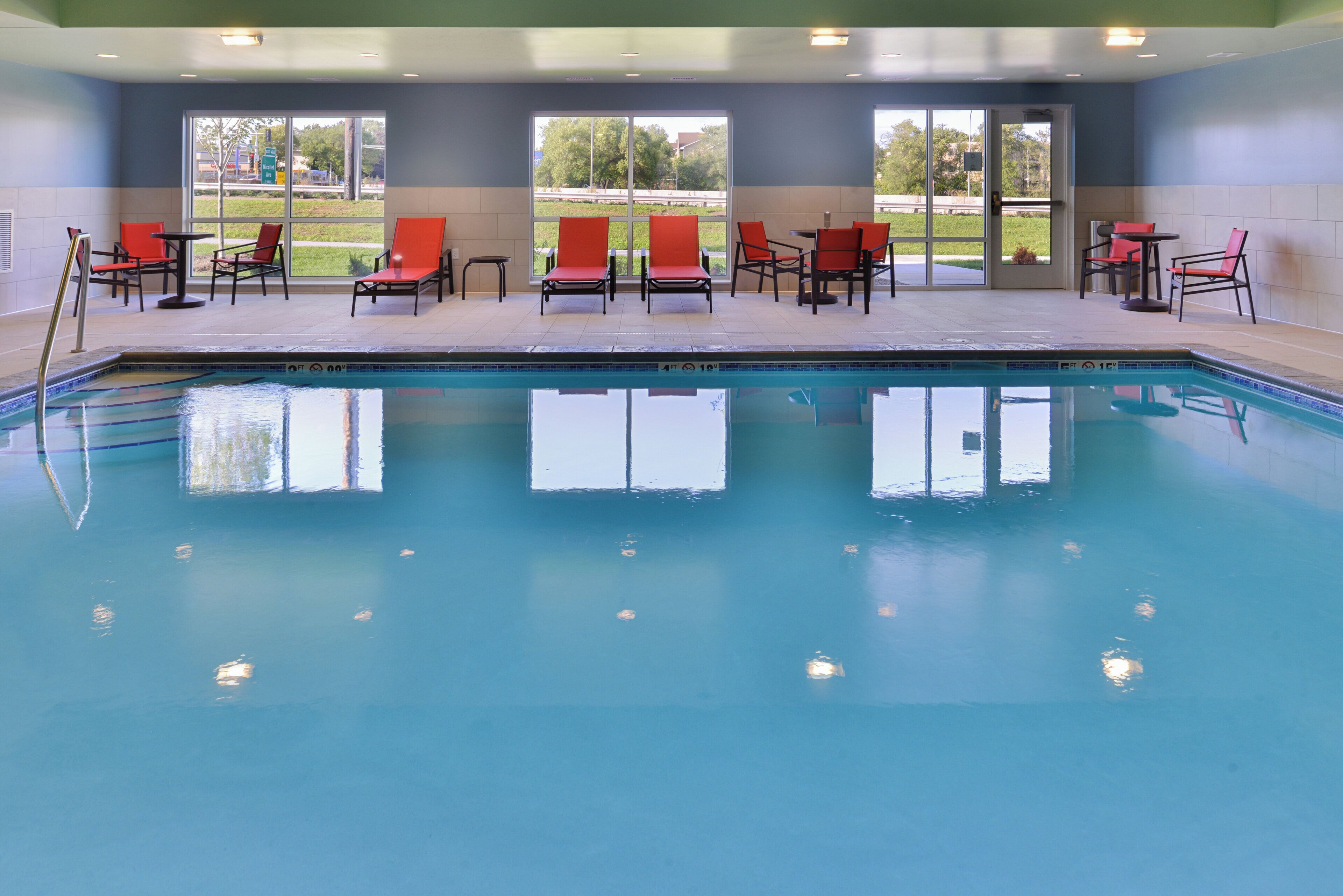 Piscina interna com água cristalina, rodeada por cadeiras e mesas vermelhas, e amplas janelas com vista para o verde.