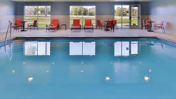 Piscina interna com água cristalina, rodeada por cadeiras e mesas vermelhas, e amplas janelas com vista para o verde.