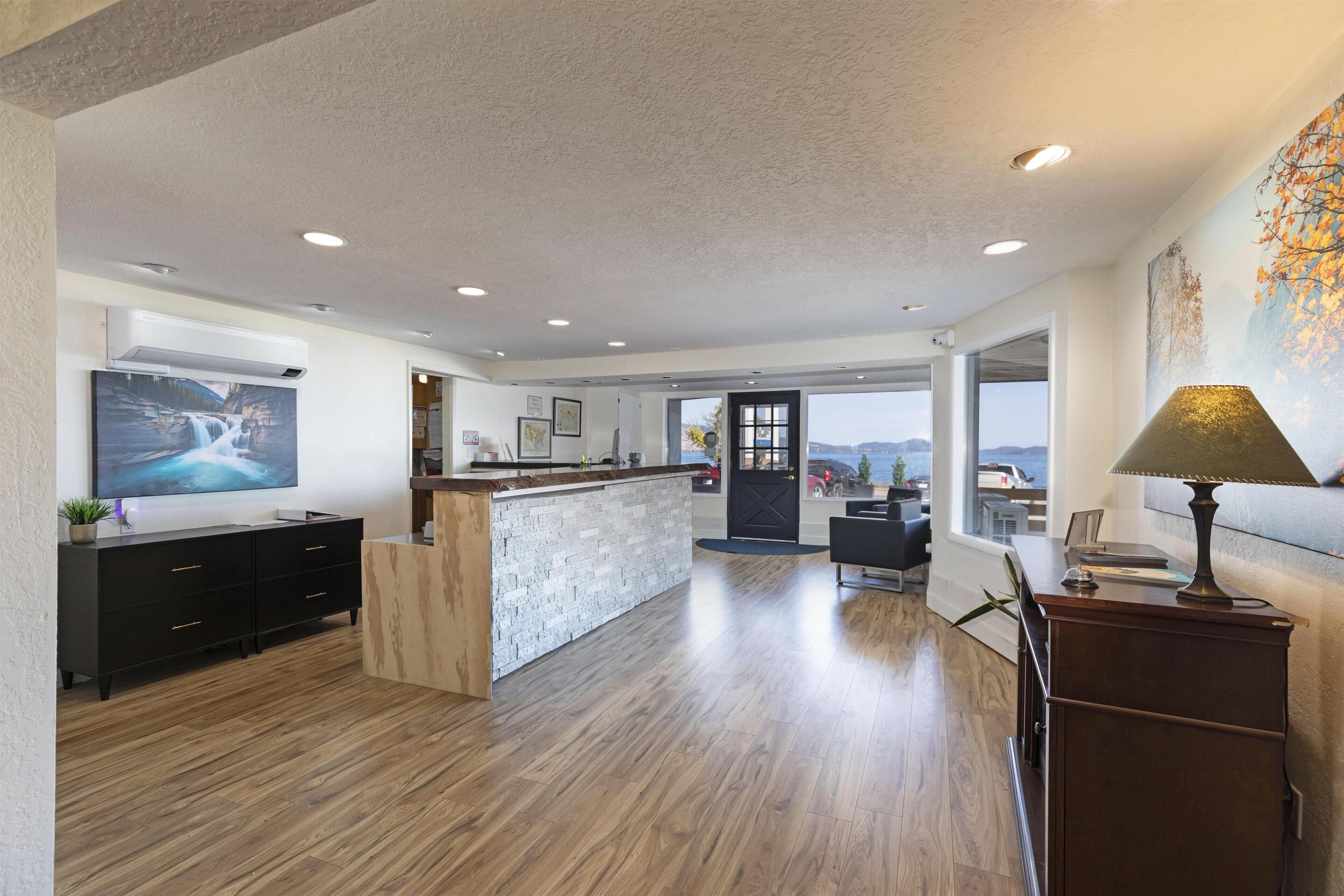 Uma sala de estar ampla com piso de madeira, uma ilha central com painel de tijolos brancos, um sofá preto e um abajur sobre uma mesa de madeira.