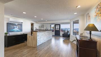 Uma sala de estar ampla com piso de madeira, uma ilha central com painel de tijolos brancos, um sofá preto e um abajur sobre uma mesa de madeira.