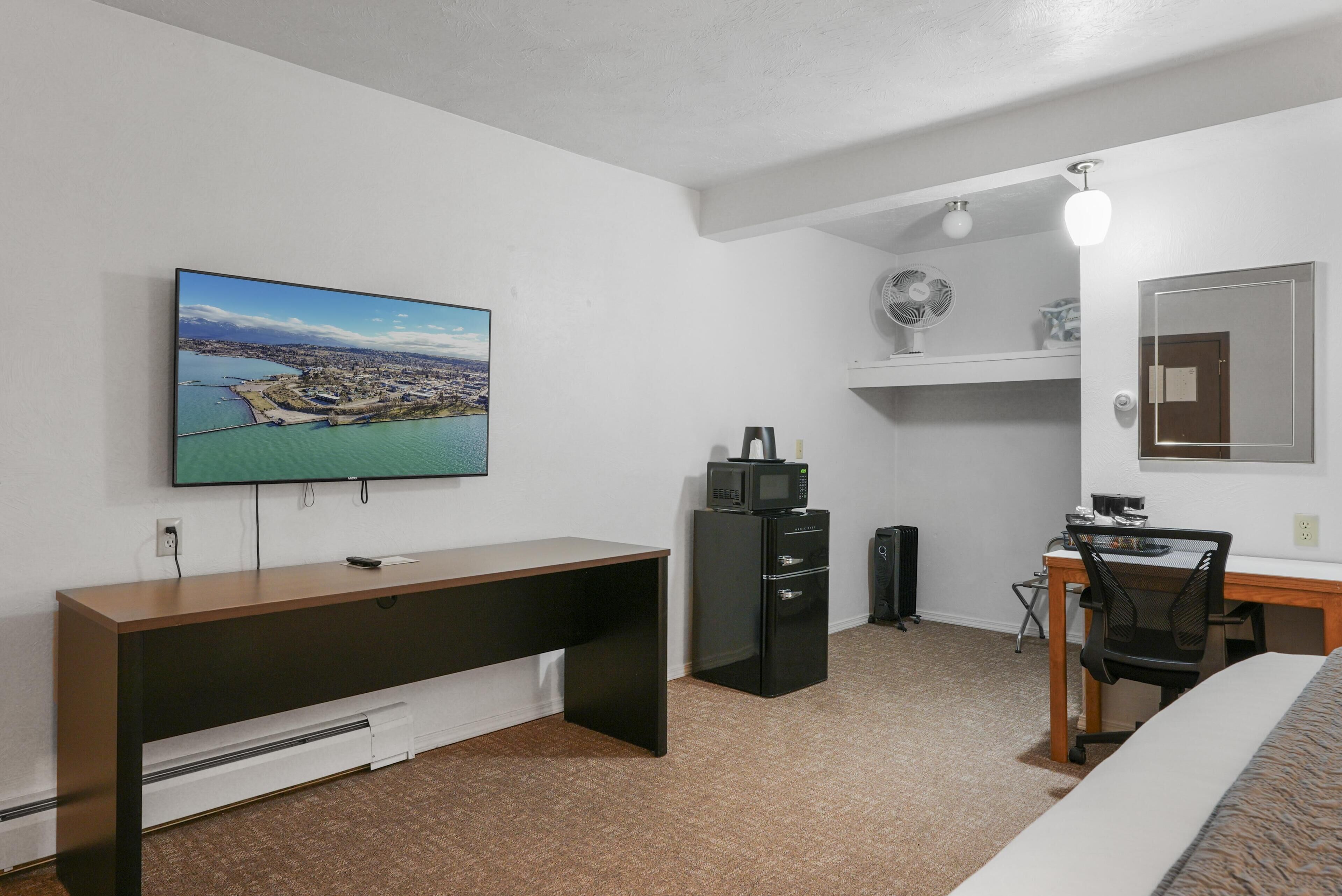 Quarto de hotel com uma TV grande na parede, uma escrivaninha com um computador, uma cadeira e uma cama.