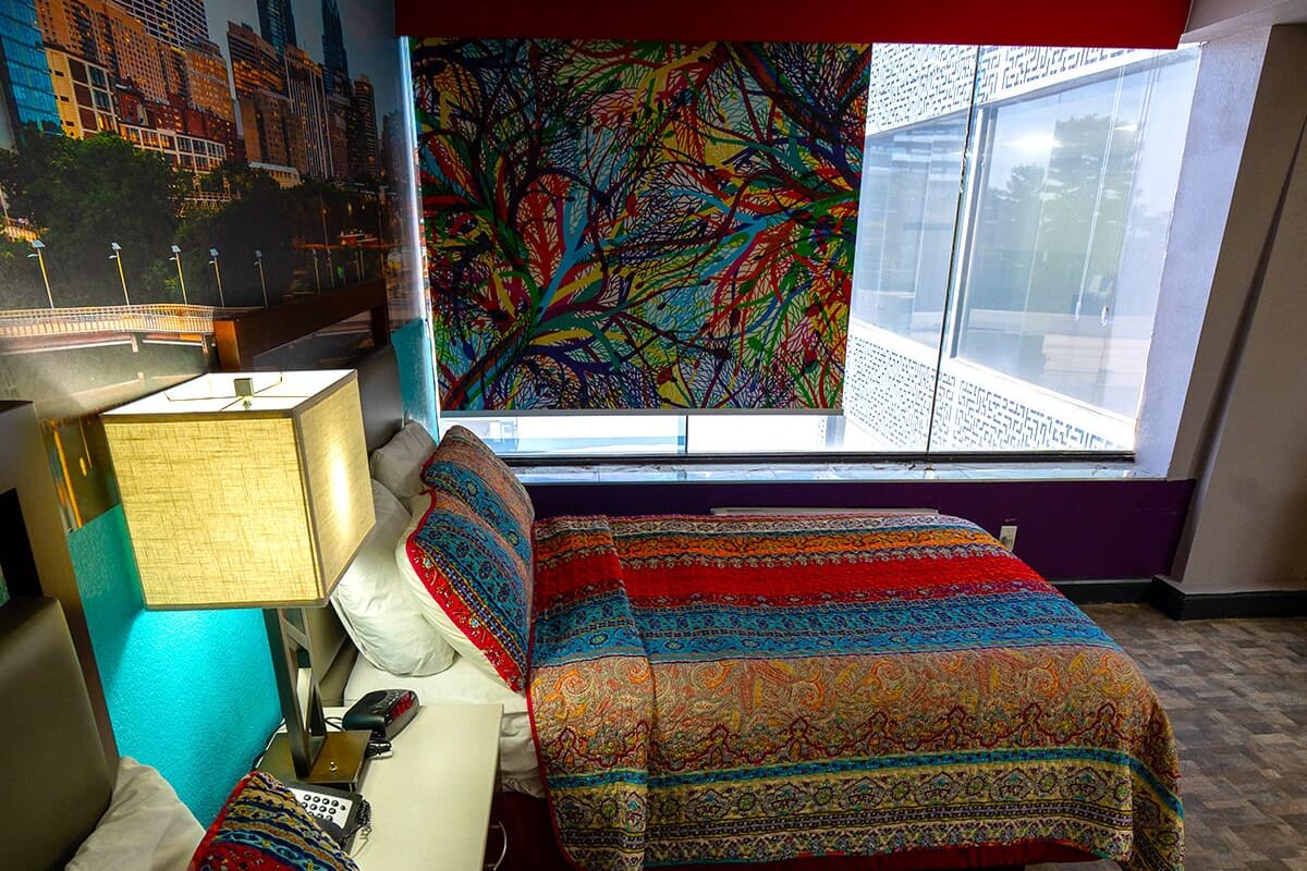 Quarto de hotel com duas camas, uma escrivaninha, uma luminária e uma janela grande com vista para prédios.