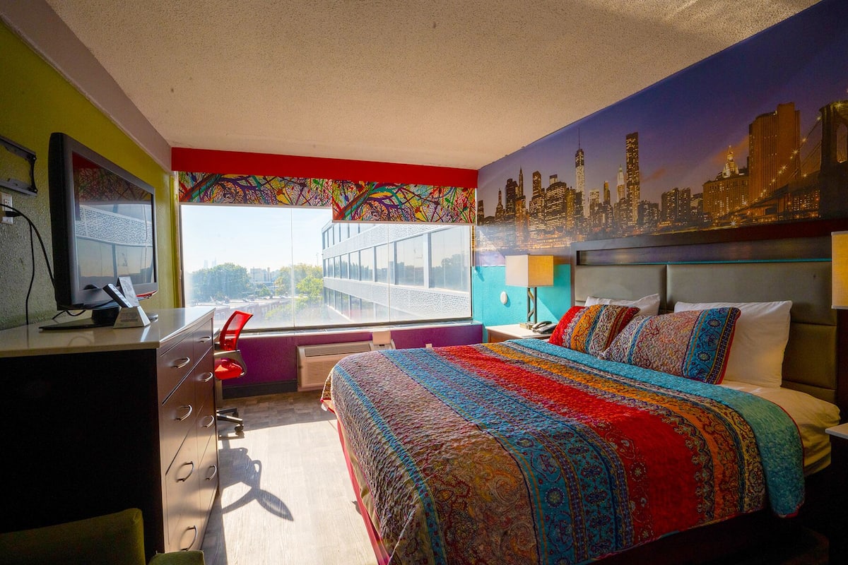 Quarto de hotel com uma cama grande, uma escrivaninha com computador, uma cadeira e uma janela com vista para a cidade.