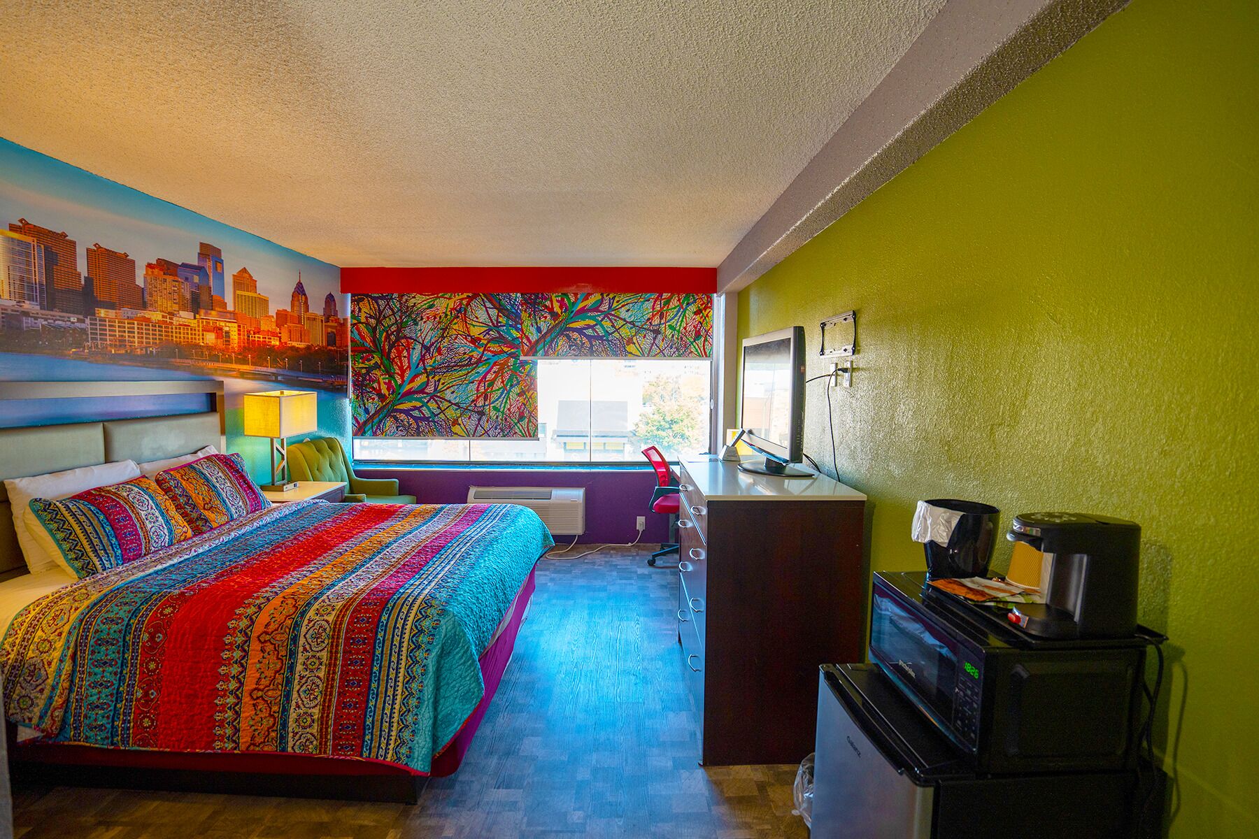 Quarto de hotel com cama, escrivaninha, computador, micro-ondas e uma grande janela.