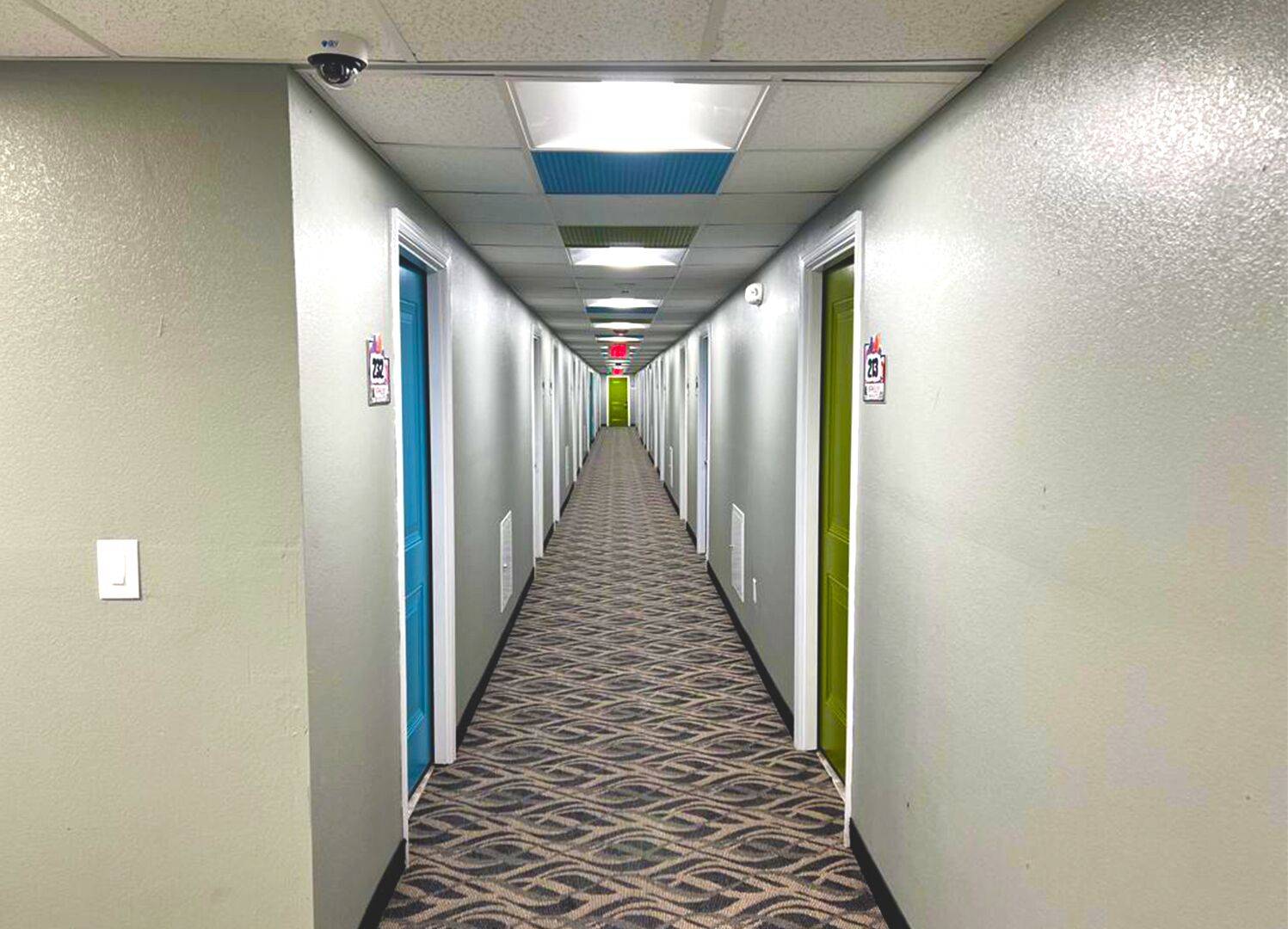 Corredor de hotel com piso coberto por carpete, portas em ambos os lados e uma câmera de segurança no teto.