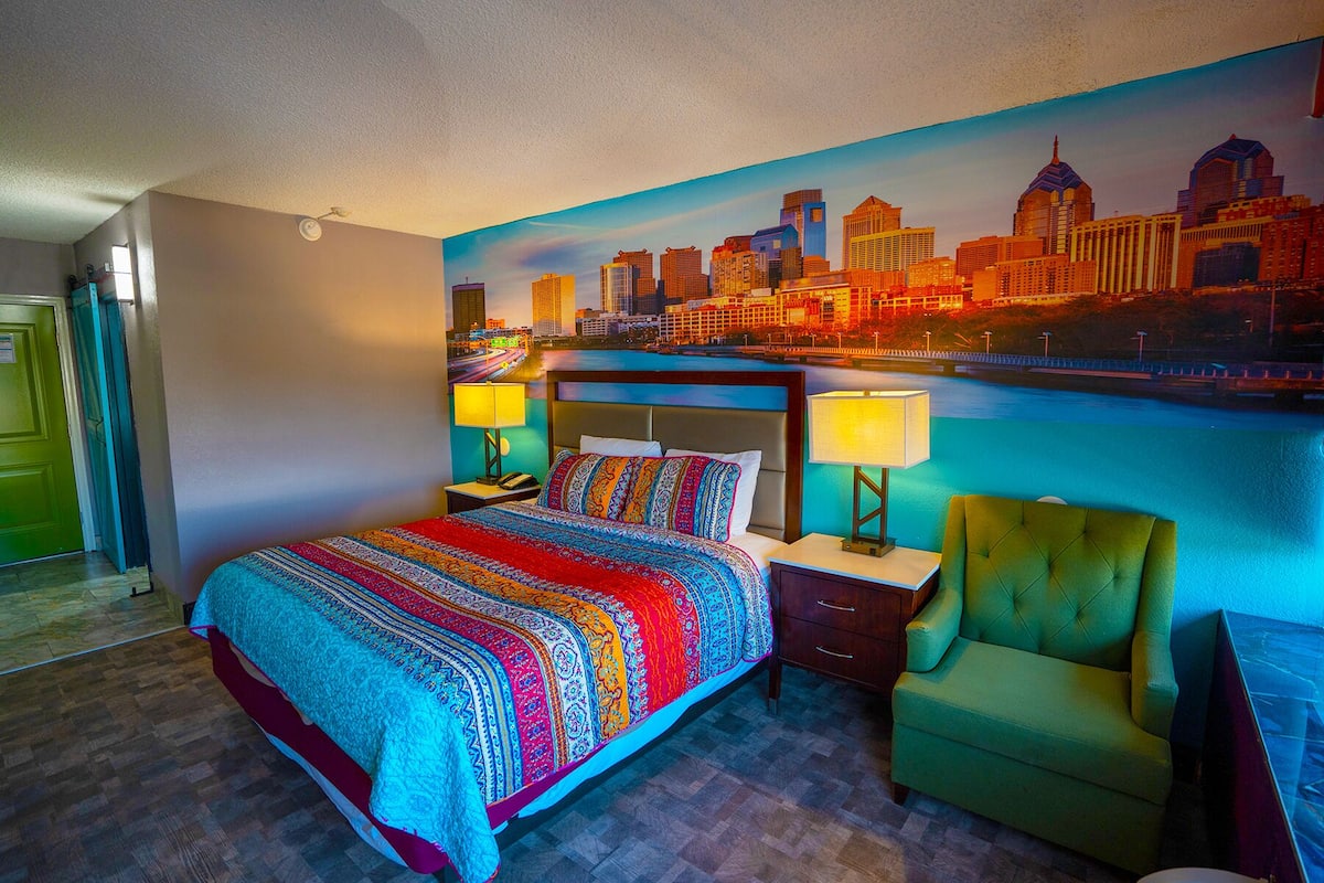 Quarto de hotel com uma cama, duas mesas de cabeceira com luminárias, uma poltrona verde e um mural com vista para a cidade na parede.