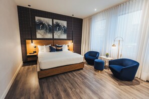 Quarto de hotel moderno com uma cama grande, duas poltronas, uma mesa pequena e arte abstrata na parede.