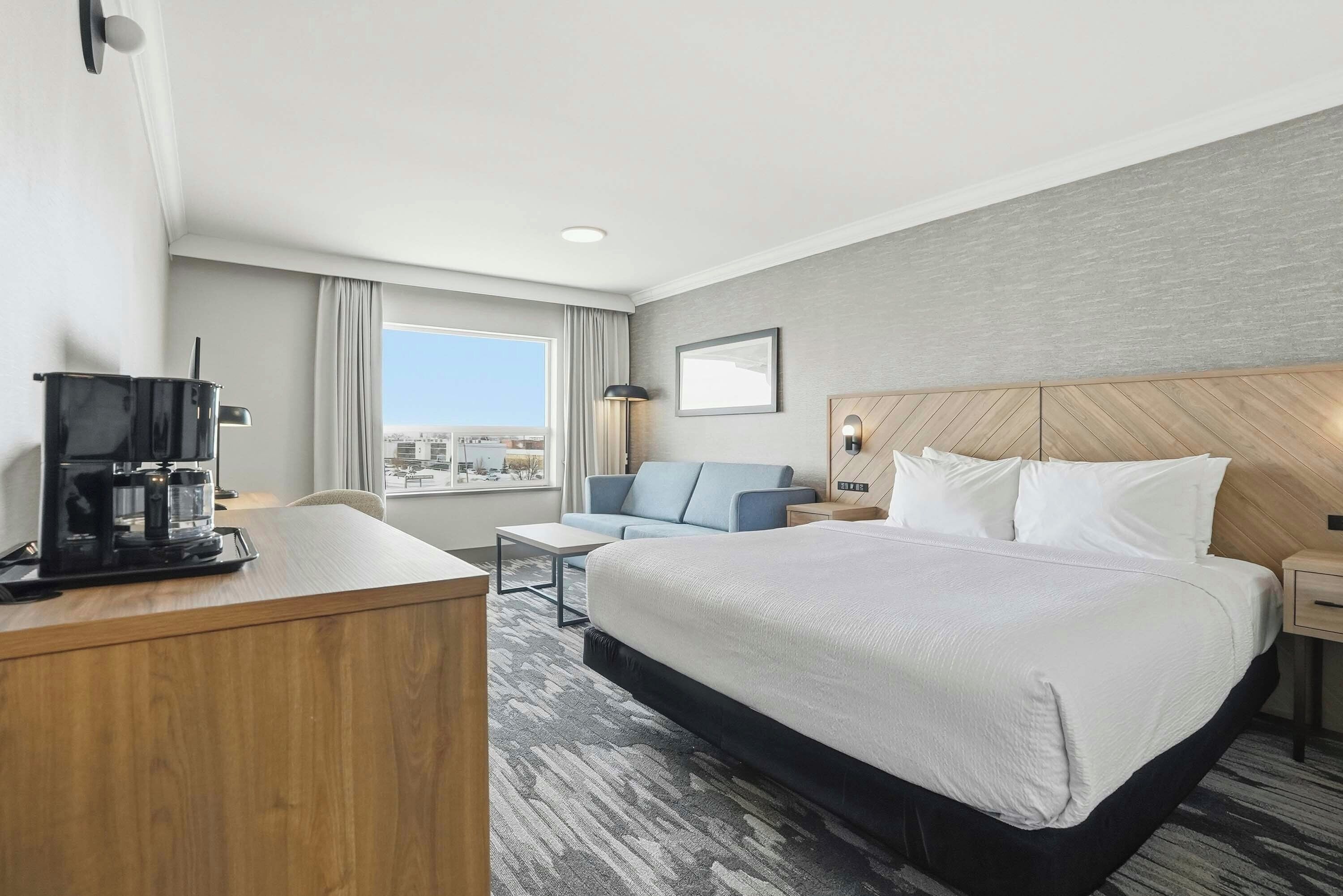 Quarto de hotel com uma cama grande, uma escrivaninha com cafeteira, um sofá e vista para a cidade.