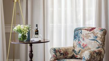 Quarto com poltrona estampada, mesa redonda com um vaso de flores, uma garrafa e um abajur.