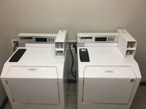 Duas máquinas de lavar roupa brancas de abertura frontal, com painéis de controlo, um cabo de alimentação e um painel de controlo fixado à parede.