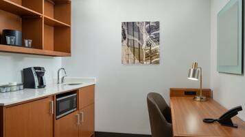 Quarto de hotel com cozinha compacta, escrivaninha, cadeira, abajur e uma obra de arte emoldurada na parede.