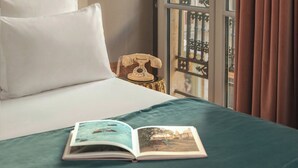 Quarto de hotel com cama, um livro, telefone antigo, luminária pendente e vista através de uma porta de vidro.