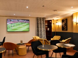 Quarto com uma televisão grande exibindo uma partida de futebol, cadeiras amarelas, uma mesa redonda e uma estante de livros.