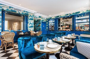 Restaurante em tons de azul, com sofás de veludo, mesas redondas, cadeiras de vime e um bar visível com prateleiras cheias de garrafas e copos.