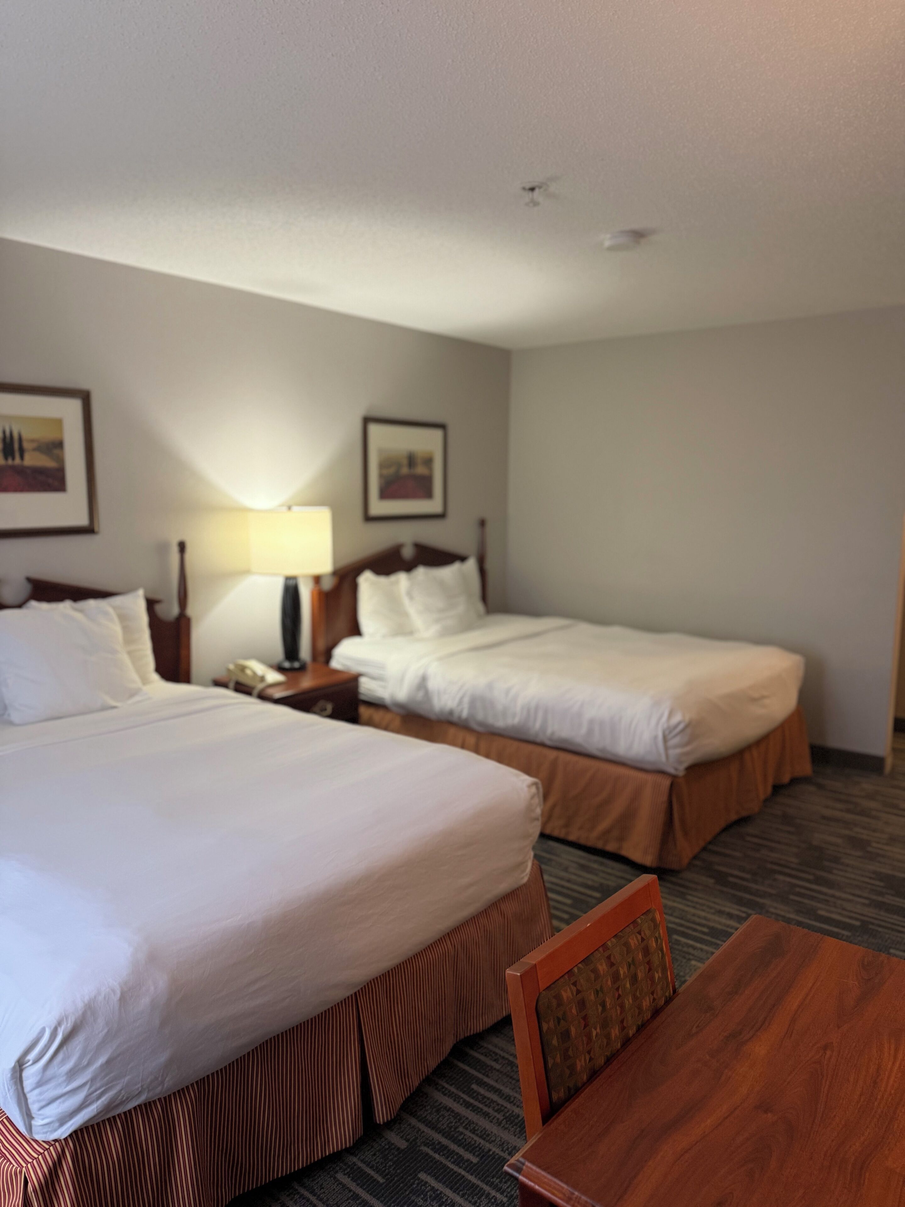 Quarto de hotel com duas camas de solteiro, uma mesa de cabeceira de madeira com abajur, uma pequena mesa e uma cadeira.