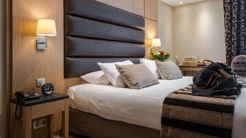 Quarto de hotel com uma cama grande, mesa de cabeceira com abajur, relógio e tomada.