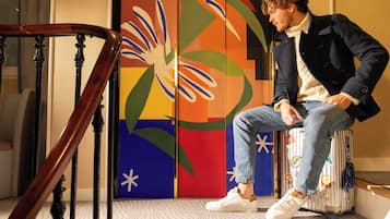 Uma pessoa sentada sobre uma mala em um quarto com um mural abstrato e colorido na parede.