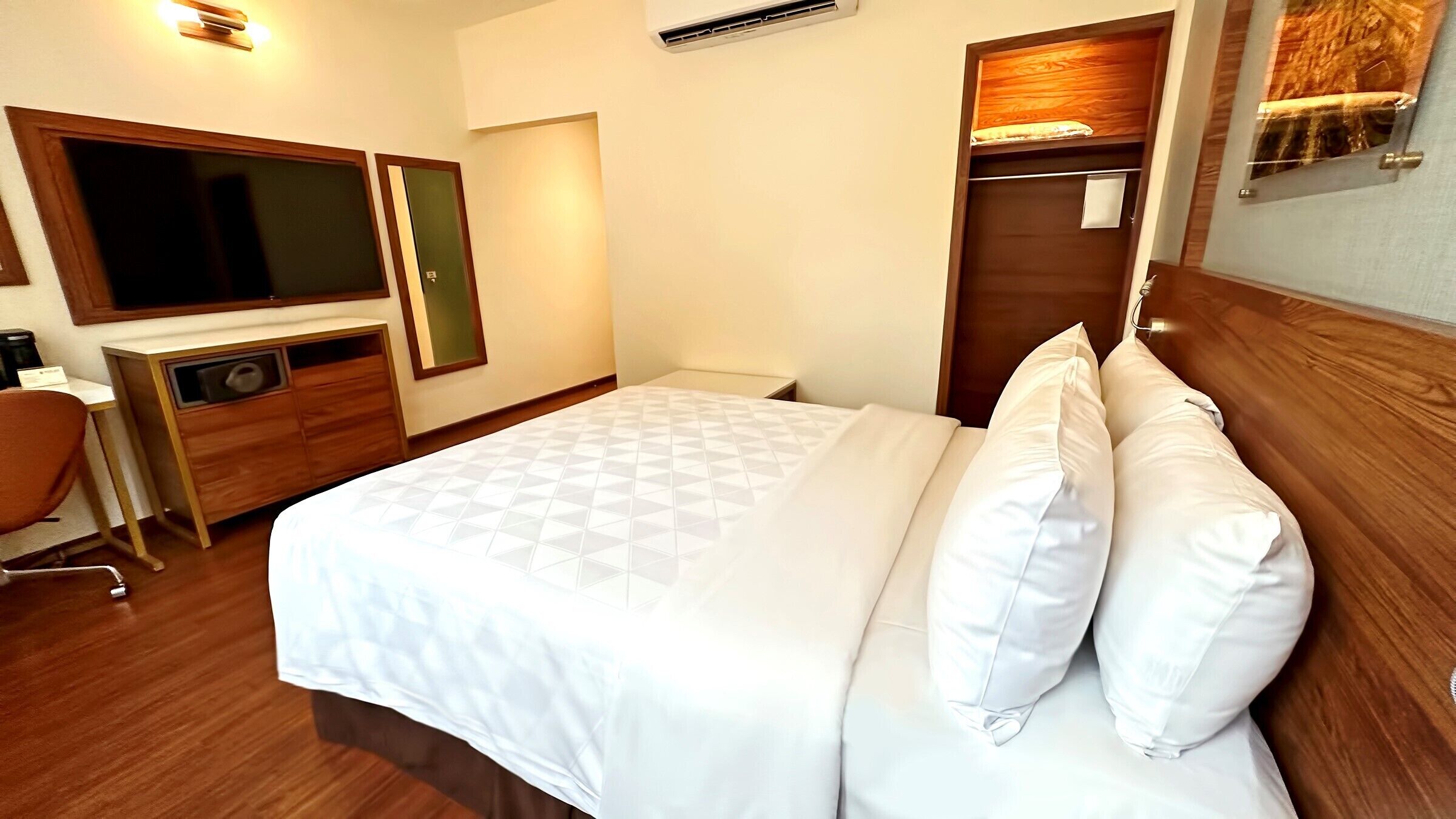 Quarto de hotel com cama, escrivaninha, cadeira, televisão e guarda-roupa.