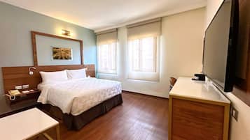 Quarto de hotel com cama, mesa de cabeceira, escrivaninha, televisão e janelas.