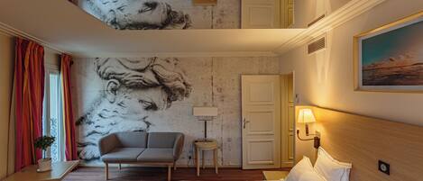 Quarto de hotel com cama, poltrona, escrivaninha, luminária e um quadro na parede.