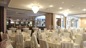 Uma sala de jantar formal com mesas redondas, toalhas brancas e cadeiras, uma área de bar com prateleiras de garrafas e lustres.