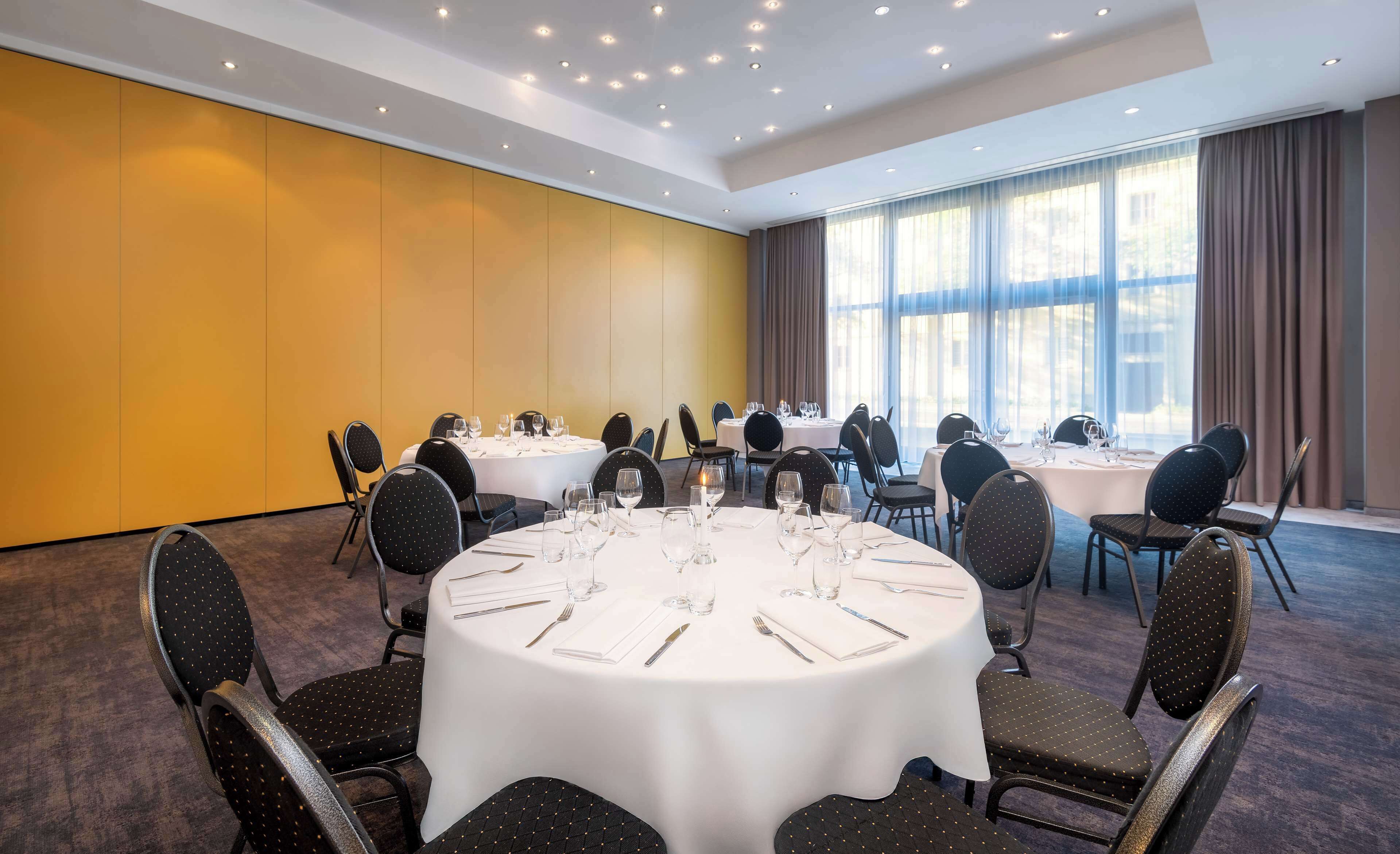 Sala de conferências com mesas redondas, cadeiras e uma grande janela com cortinas.