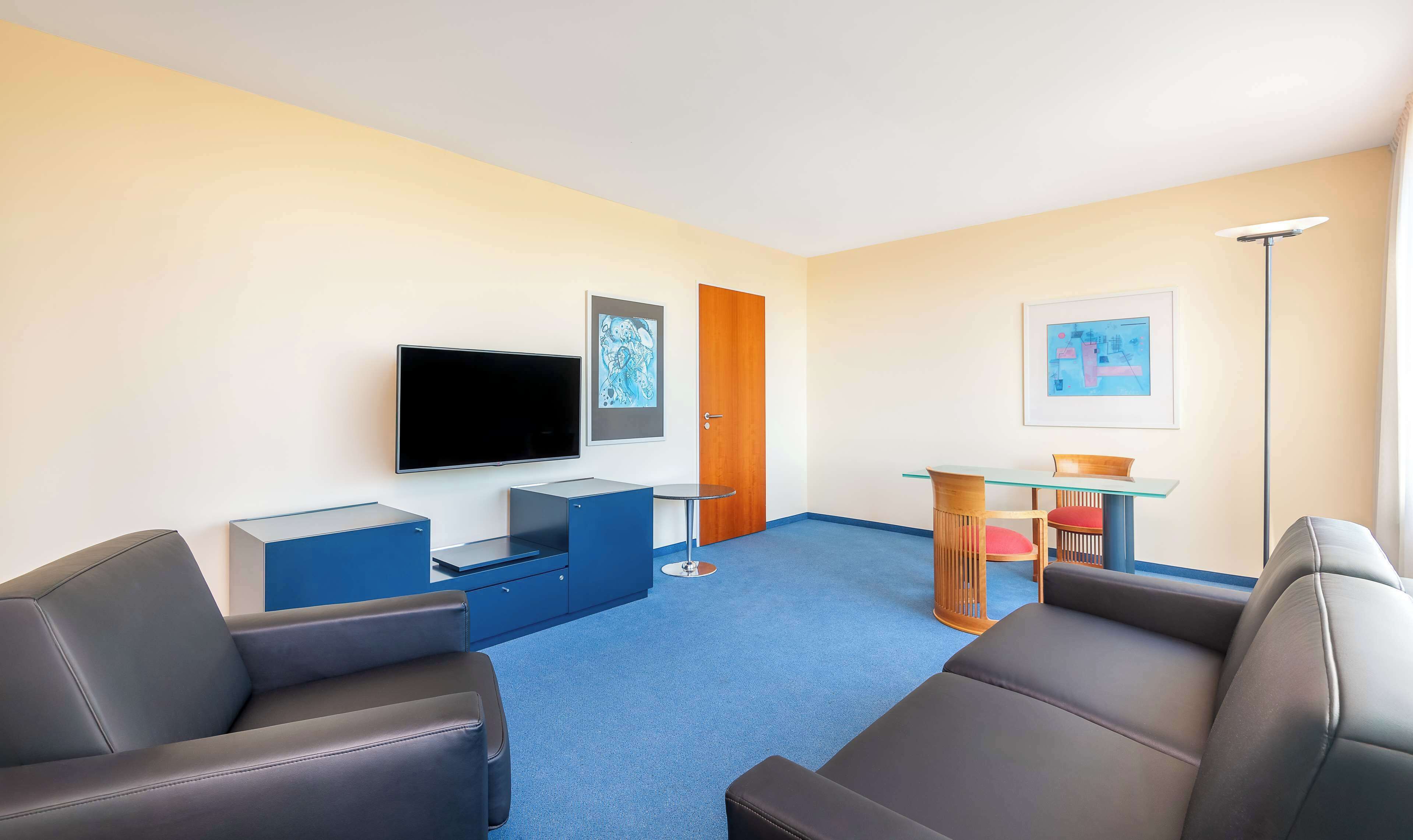 Quarto de hotel moderno com televisão de tela plana, mesa de vidro, dois sofás azuis e porta de madeira.