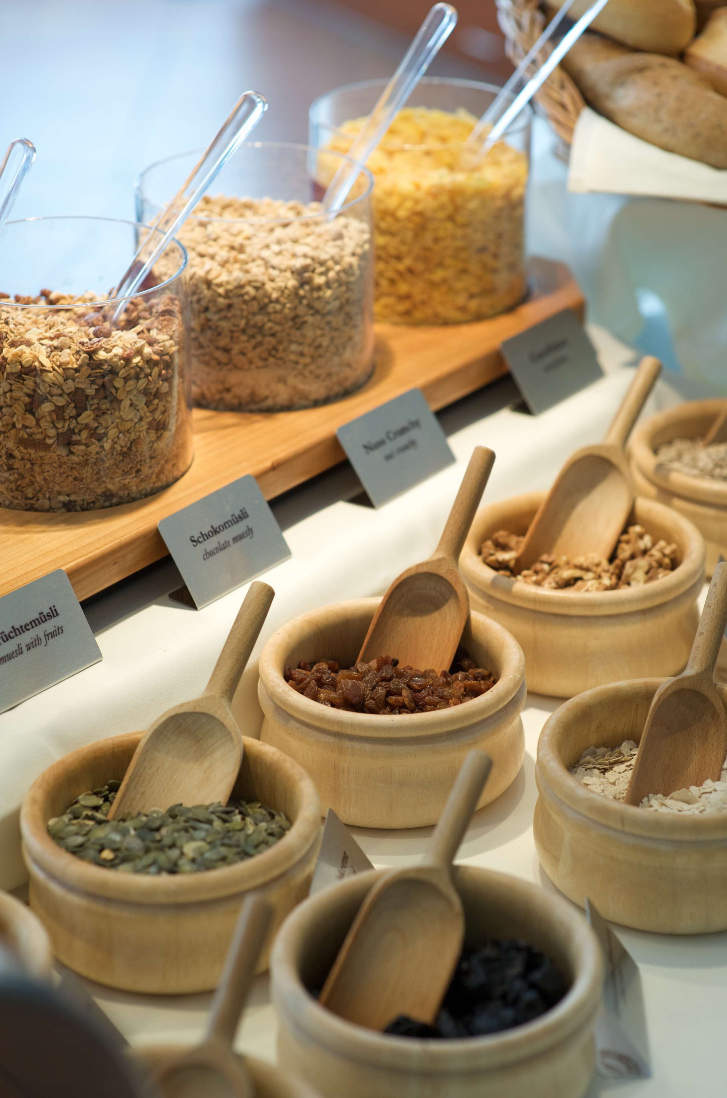 Diversos alimentos apresentados em tigelas de madeira com etiquetas, incluindo chocolate e granola.