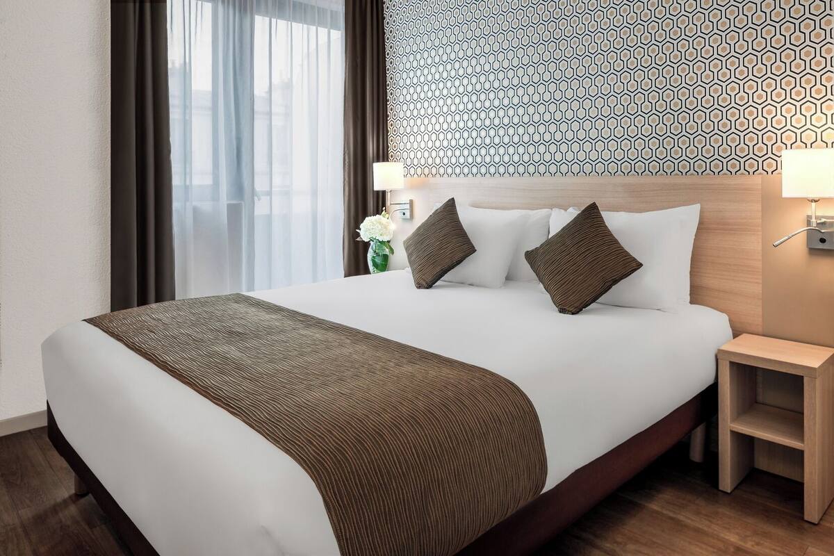 Quarto de hotel com uma cama grande, cabeceira de madeira, mesa de cabeceira, abajur e parede estampada.