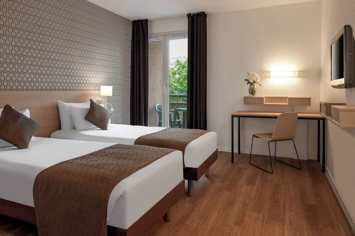 Quarto de hotel com duas camas, uma mesa, uma cadeira, televisão e varanda com vista para vegetação.