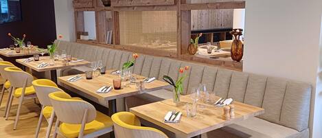 Interior de restaurante com várias mesas, cadeiras acolchoadas amarelas e um painel de madeira com plantas.