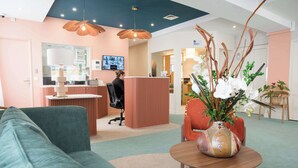 Um lobby moderno de hotel com um sofá azul-turquesa, um balcão de recepção de madeira e um vaso decorativo com flores brancas.