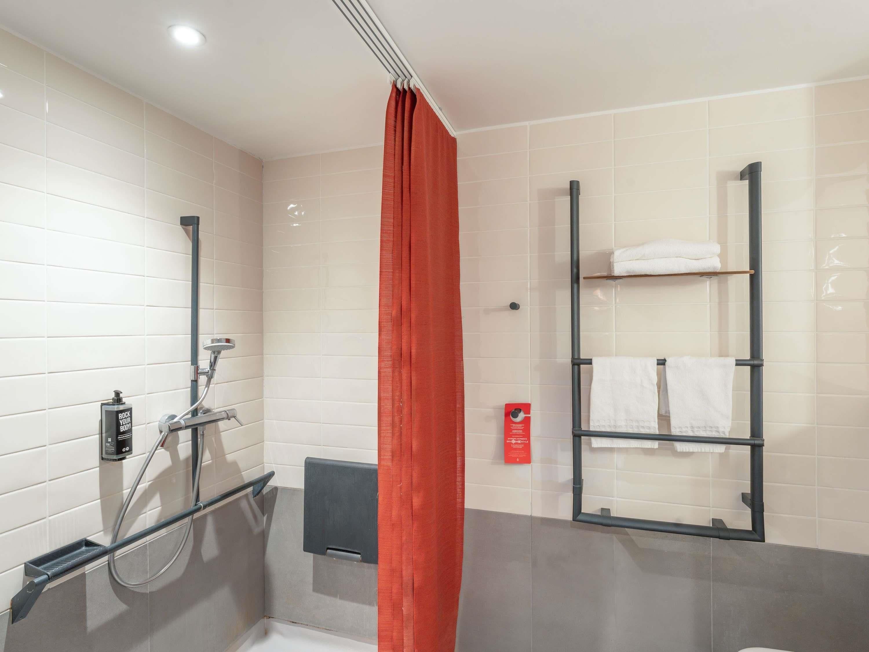 Casa de banho moderna com azulejos brancos e cinzentos, chuveiro com porta de vidro, porta-toalhas com toalhas dobradas e dispensador de sabonete.