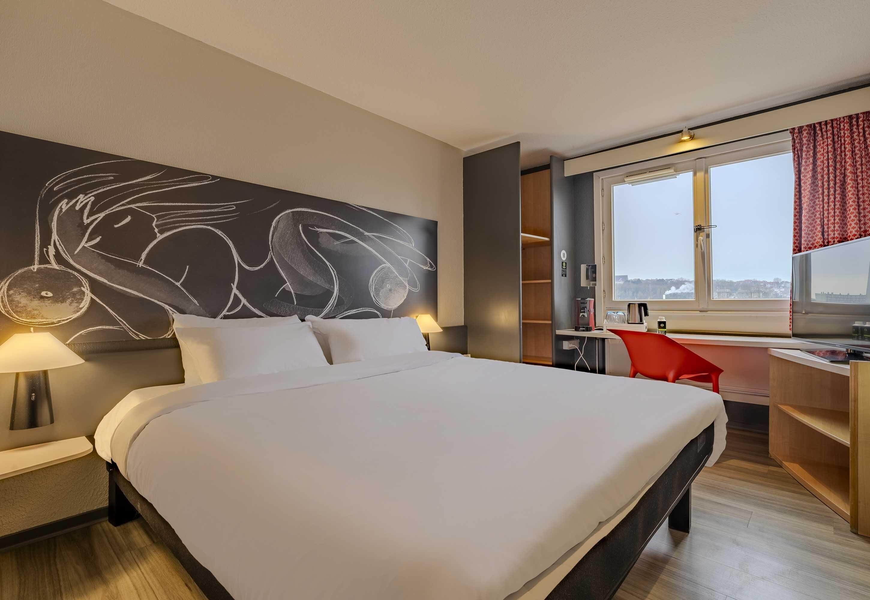 Quarto de hotel moderno com uma cama grande, uma escrivaninha, uma cadeira e vista para a cidade.