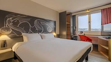 Quarto de hotel moderno com uma cama grande, uma escrivaninha, uma cadeira e vista para a cidade.