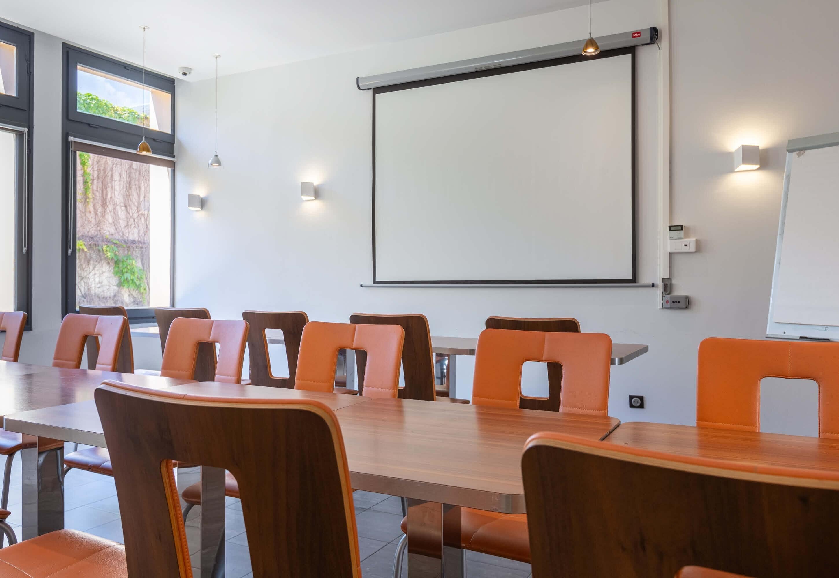 Uma sala de conferência com uma mesa grande, cadeiras laranja, uma tela de projetor e um quadro branco.