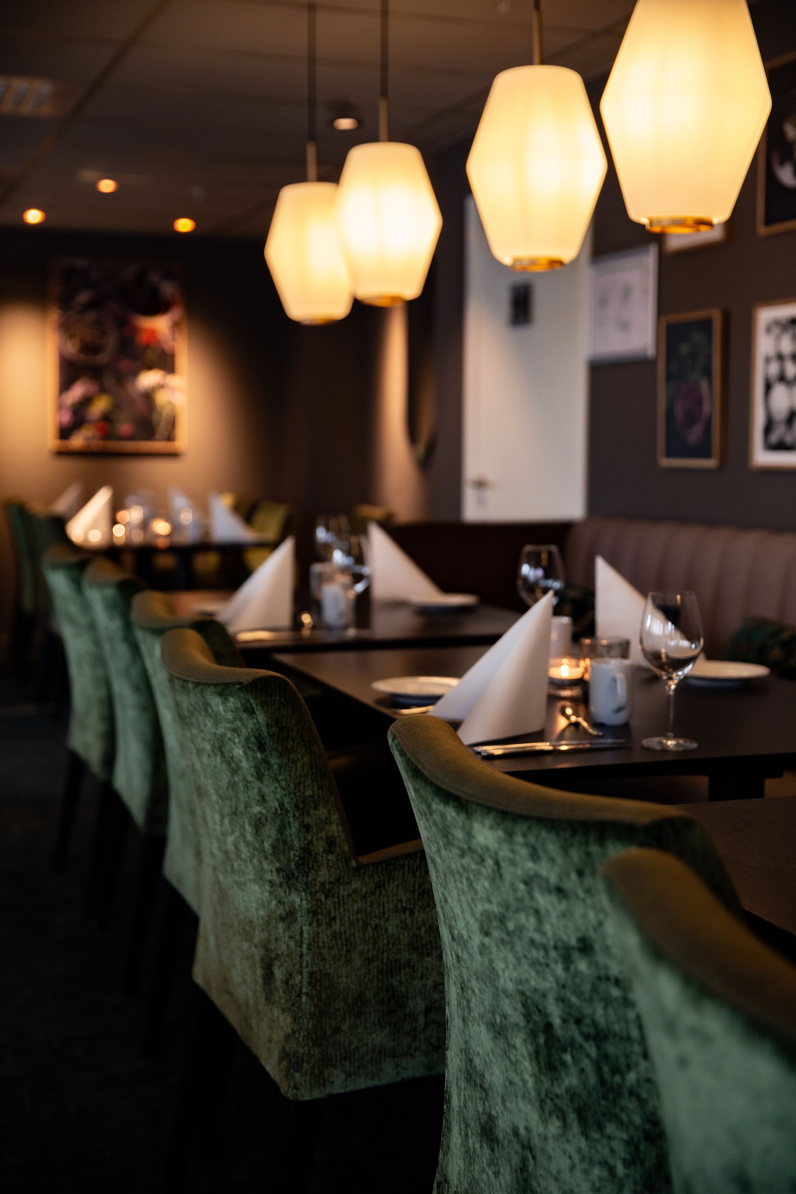 Restaurante com cadeiras estofadas em verde, mesas postas com guardanapos brancos, copos de vinho e uma vela sobre a mesa.