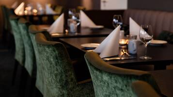 Restaurante com cadeiras estofadas em verde, mesas postas com guardanapos brancos, copos de vinho e uma vela sobre a mesa.