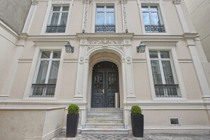 Um edifício com entrada central, ladeada por duas janelas com grades decorativas, e fachada simétrica com janelas e molduras ornamentais regularmente distribuídas.