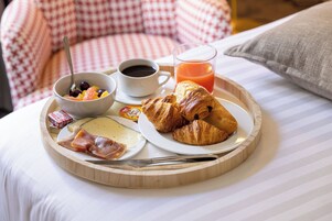 Bandeja de café da manhã com croissants, queijo, presunto e um copo de suco de laranja sobre uma cama com roupa de cama listrada.