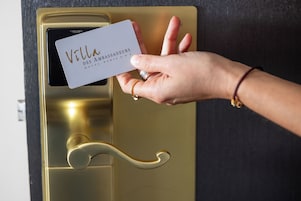 Mão segurando um cartão com a inscrição "Villa Des Ambassadeurs HOTEL PARIS", em frente a uma maçaneta dourada.