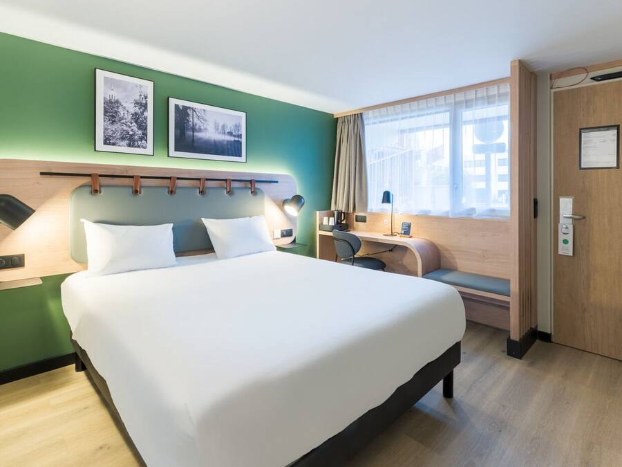 Quarto de hotel com uma cama grande, uma escrivaninha, uma cadeira, uma janela com cortinas e uma porta.