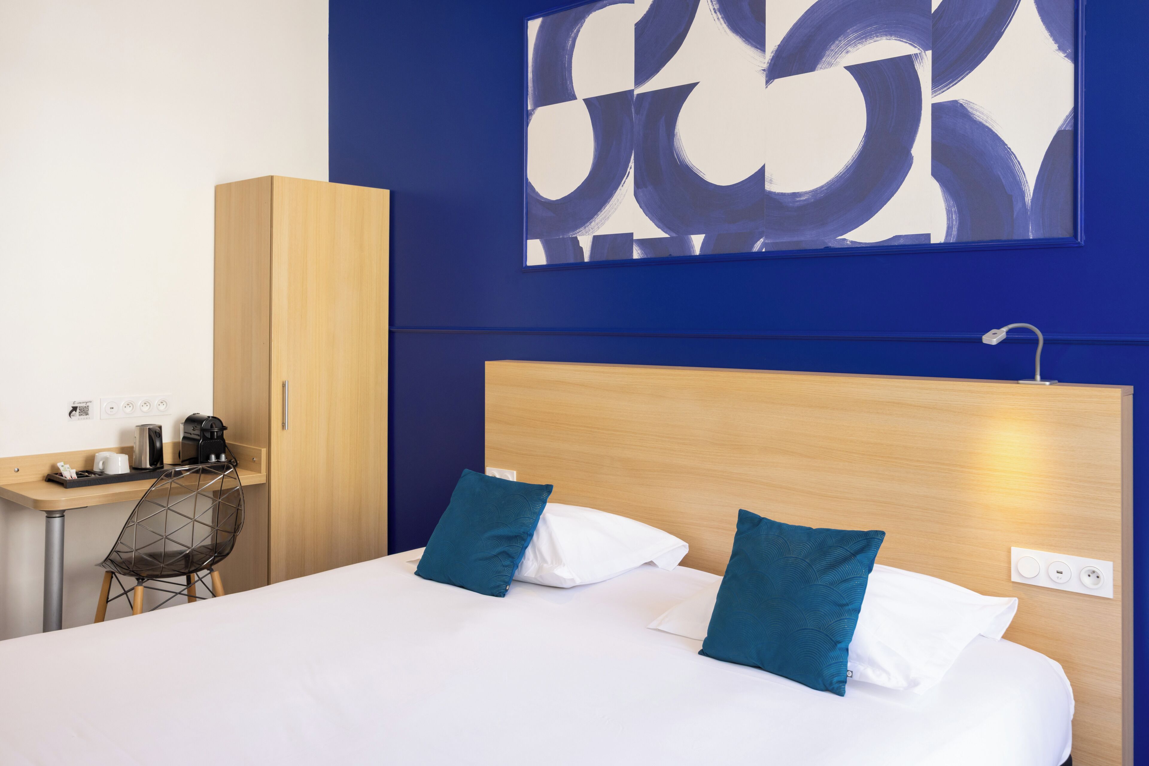 Quarto de hotel com cama, duas almofadas, escrivaninha com cadeira, cafeteira e uma obra de arte na parede.