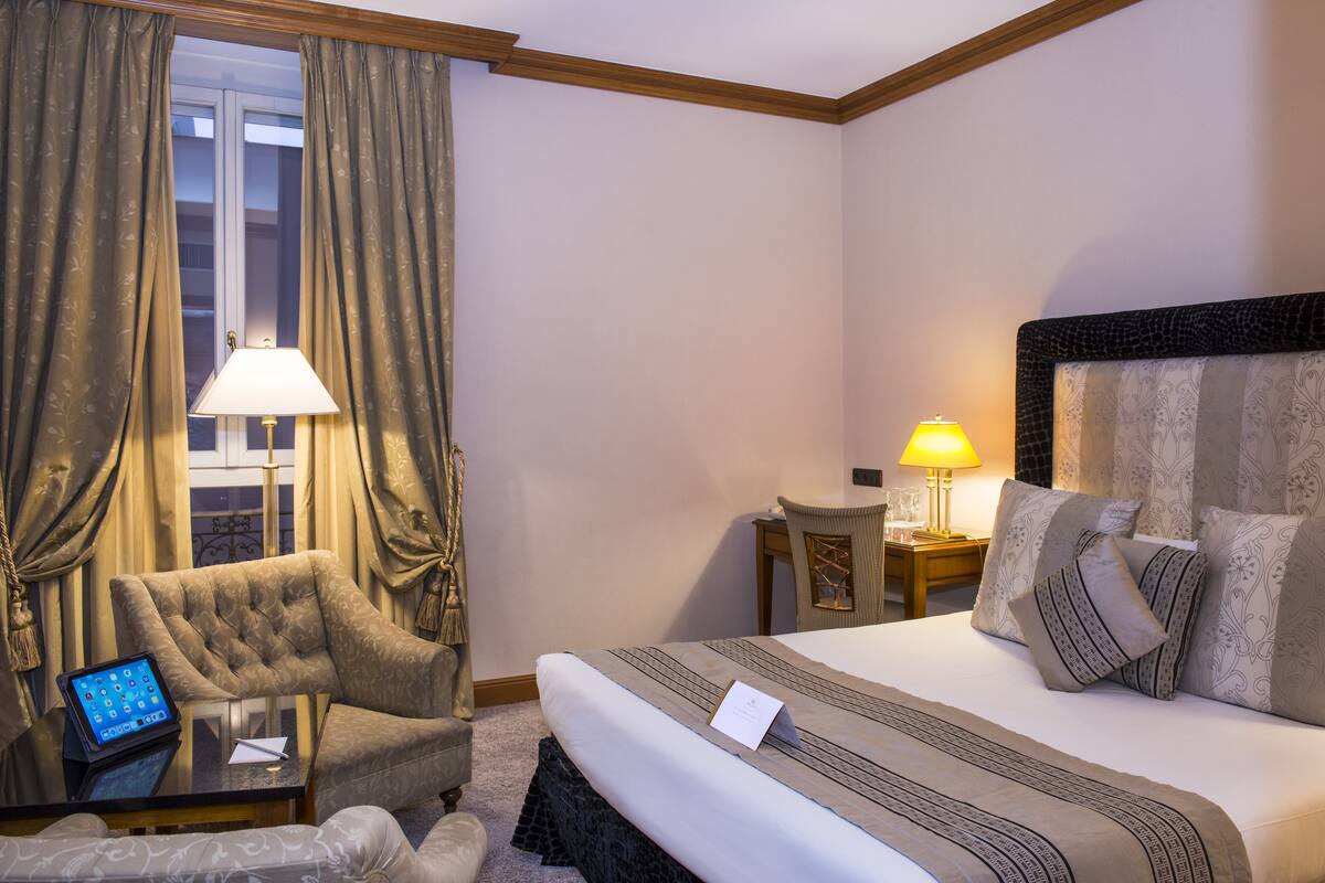 Quarto de hotel com uma cama, uma cadeira, uma escrivaninha com abajur e uma janela com cortinas.