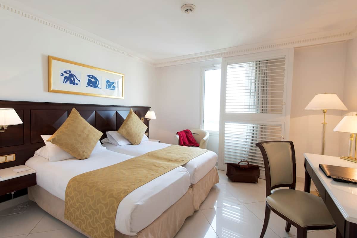 Quarto de hotel com uma cama grande, duas mesas de cabeceira, uma escrivaninha e uma cadeira. Há uma janela com persianas, um quadro na parede e uma luminária na escrivaninha.